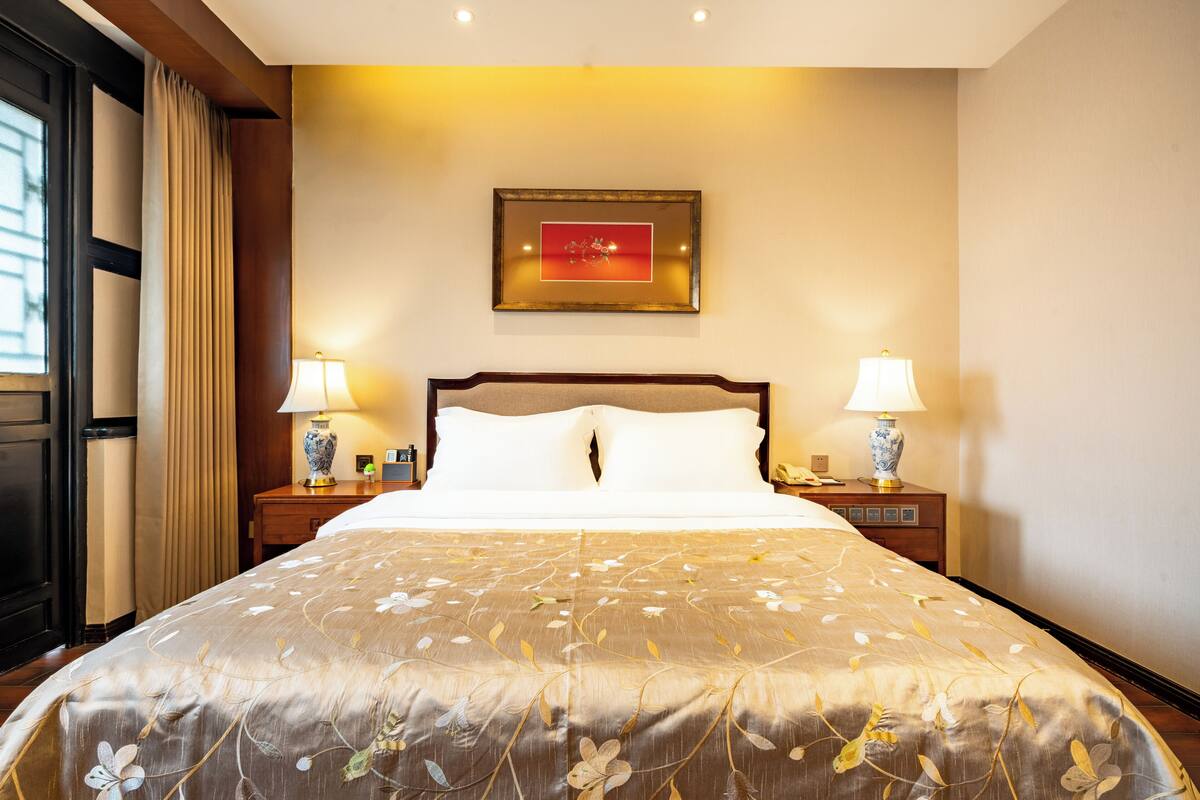 Quarto de hotel com uma cama grande, duas mesas de cabeceira com abajures, um quadro na parede e uma janela com cortinas.