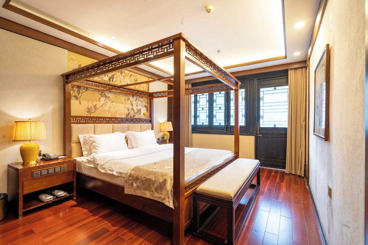 Uma cama com dossel de madeira e cabeceira estampada com flores, uma mesa de cabeceira com luminária e um banquinho aos pés da cama.