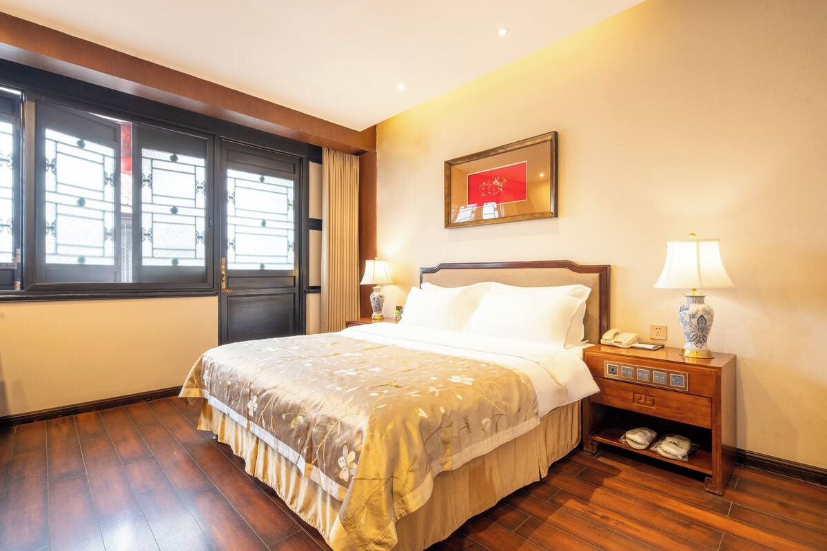 Quarto de hotel com uma cama grande, uma mesa de cabeceira com abajur, um quadro pendurado na parede e uma janela com cortinas.
