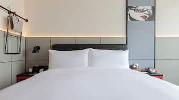 Quarto de hotel com uma cama grande, uma mesa de cabeceira com abajur, uma mesinha com um globo e um telefone, e um porta-toalhas fixado na parede.