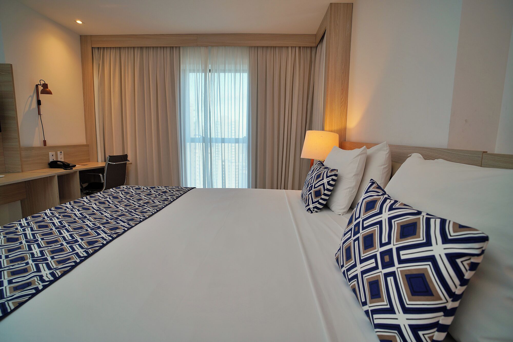 Quarto de hotel com uma cama grande, uma escrivaninha com telefone e uma luminária.