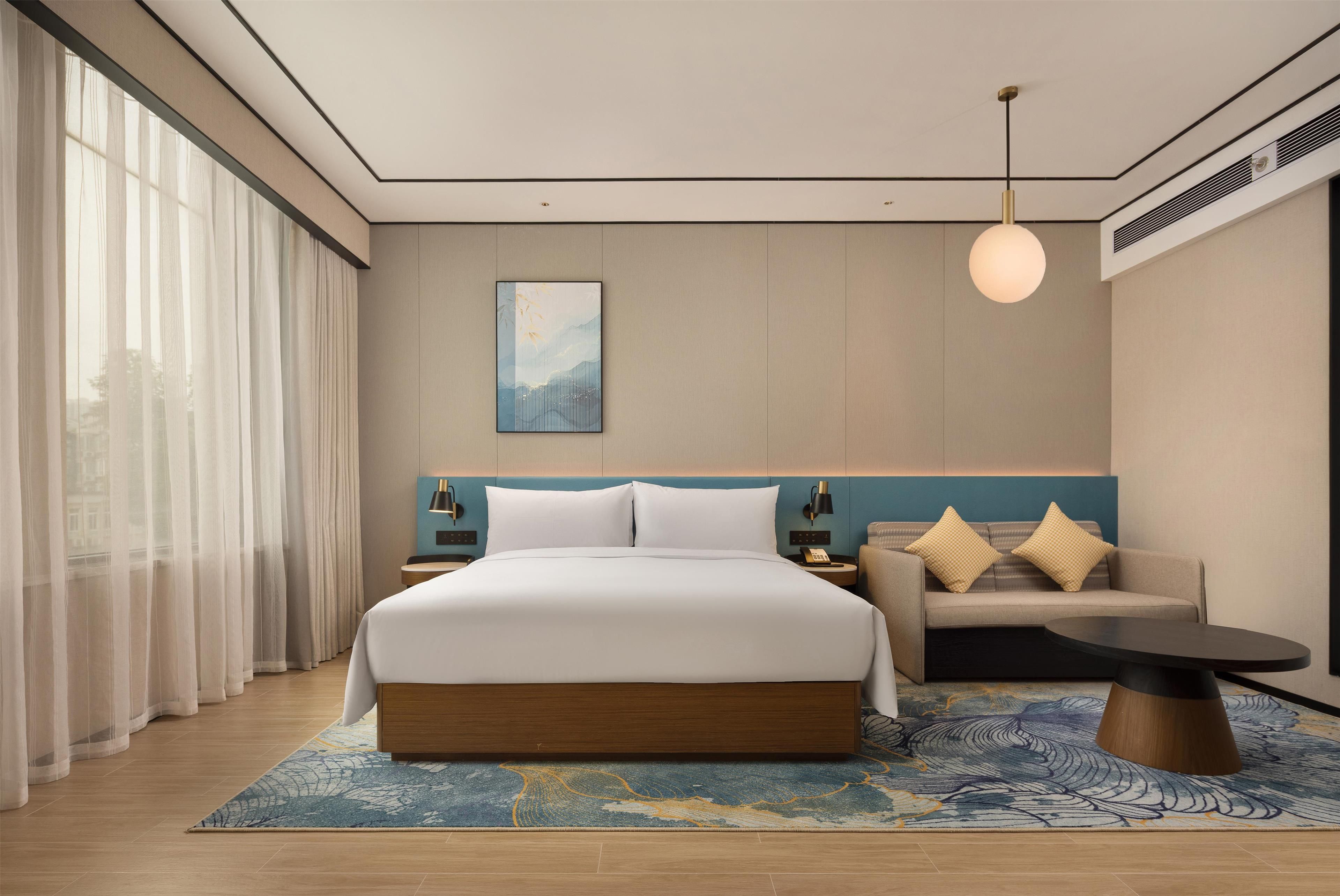 Quarto de hotel moderno com cama grande, sofá, mesa pequena e vista para a cidade.