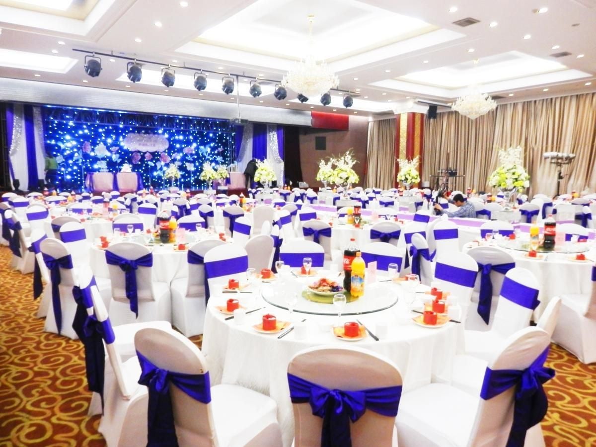 Salão de festas com mesas redondas, cadeiras brancas com fitas azuis e palco com decoração floral.