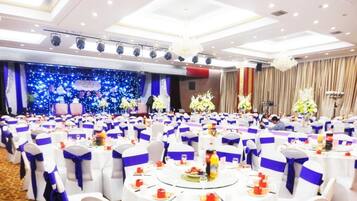 Salão de festas com mesas redondas, cadeiras brancas com fitas azuis e palco com decoração floral.