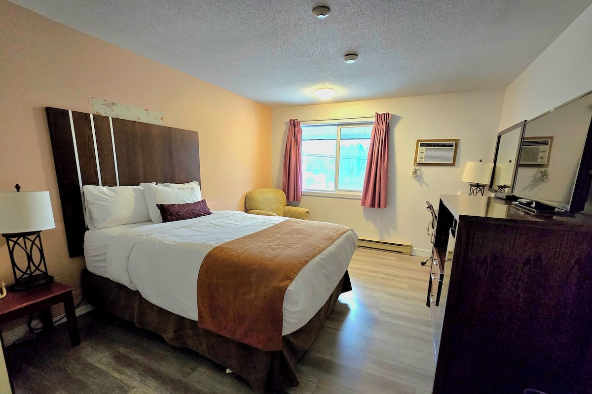 Quarto de hotel com cama, escrivaninha, cadeira, televisão e janela com cortinas.