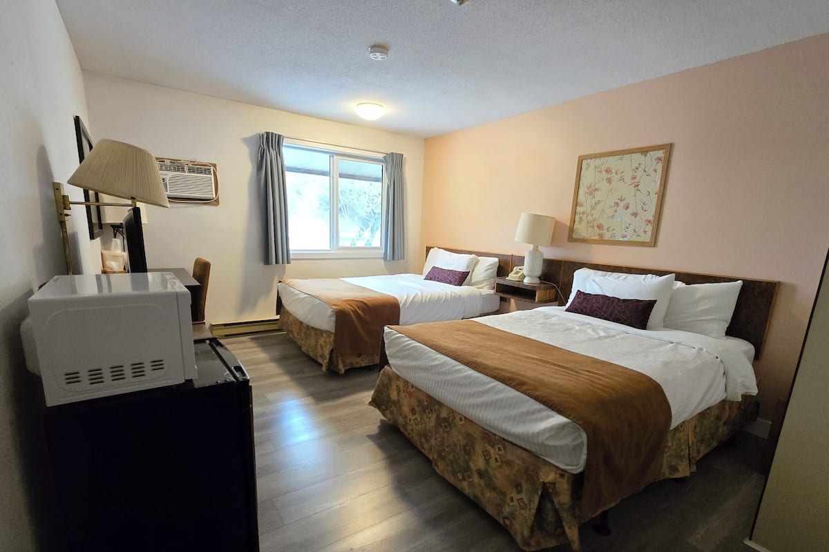 Quarto de hotel com duas camas, uma escrivaninha, um abajur, um quadro na parede e uma janela com cortinas.