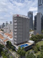 Um hotel moderno de grande porte, com fachada em linhas retas, piscina no terraço e cercado por outros edifícios contemporâneos.