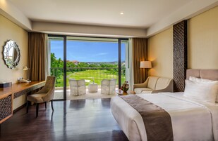 Quarto de hotel com uma cama grande, uma mesa, uma cadeira, um sofá e uma varanda com vista para uma paisagem verde.