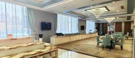 Um amplo hall de hotel com um balcão de recepção extenso, área de estar e uma grande janela que ilumina naturalmente o espaço.