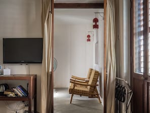 Um quarto com televisão na parede, uma cadeira de madeira, uma estante de livros e uma janela com cortinas.
