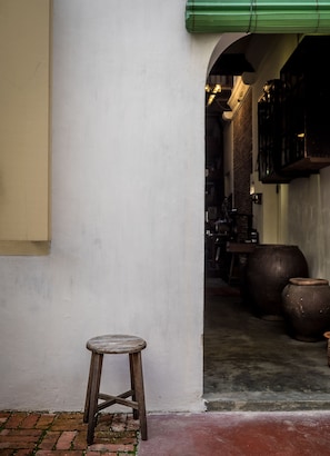 Um beco estreito com um banquinho de madeira, chão de tijolos e grandes vasos ao fundo.