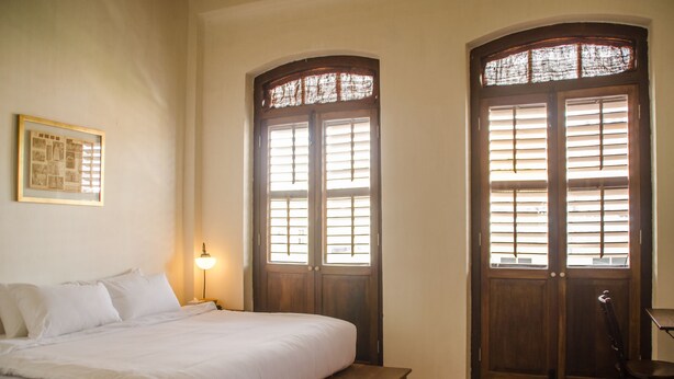 Um quarto com uma cama, um banco, duas portas de madeira com janelas e um quadro na parede.