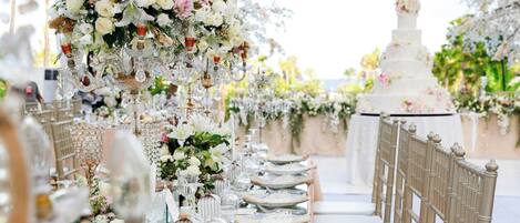 Salão de banquetes com longas mesas preparadas para um evento formal, decorado com arranjos florais e elegantes louças.