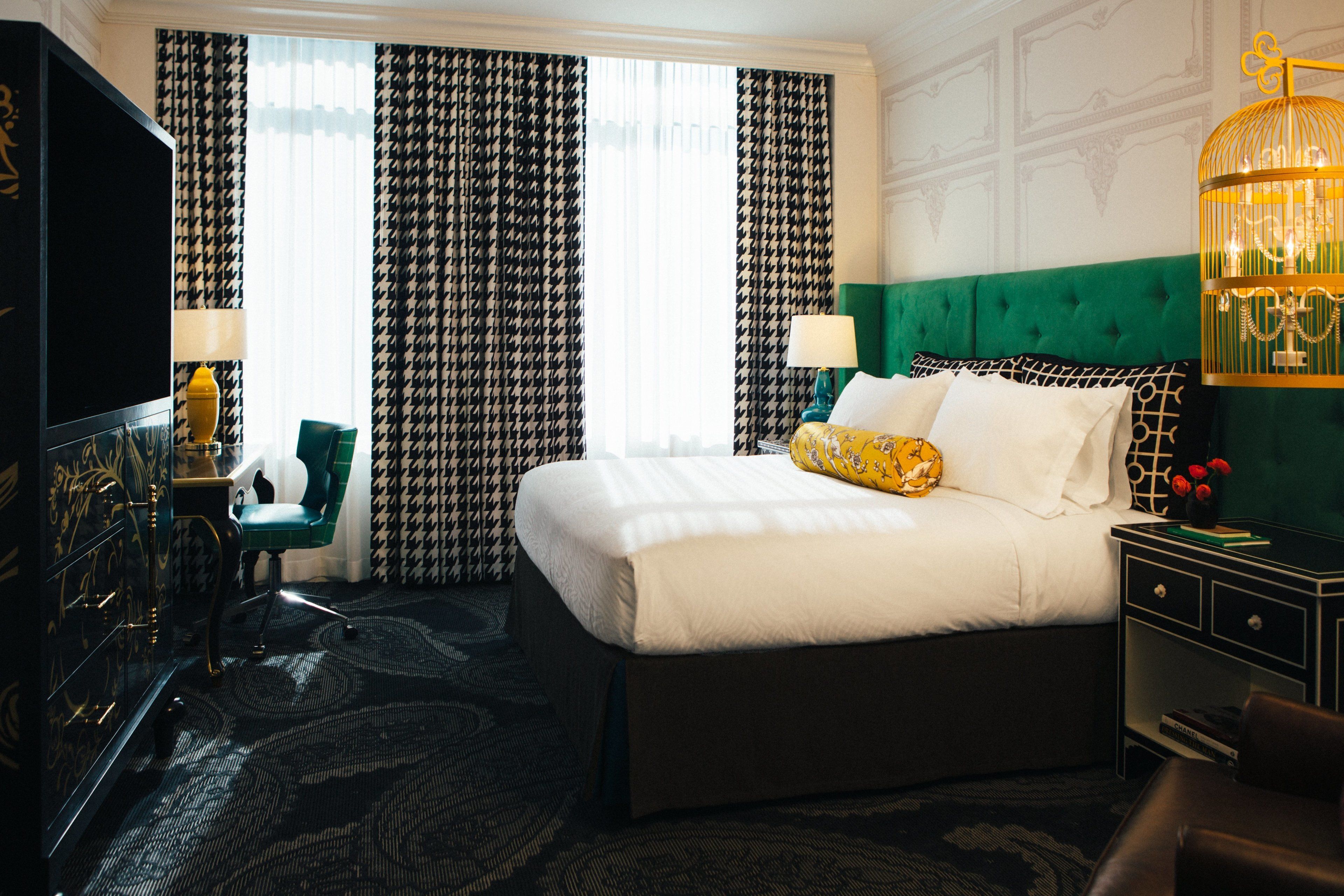 Quarto de hotel com uma cama grande, cabeceira estofada verde, uma televisão e uma gaiola decorativa.
