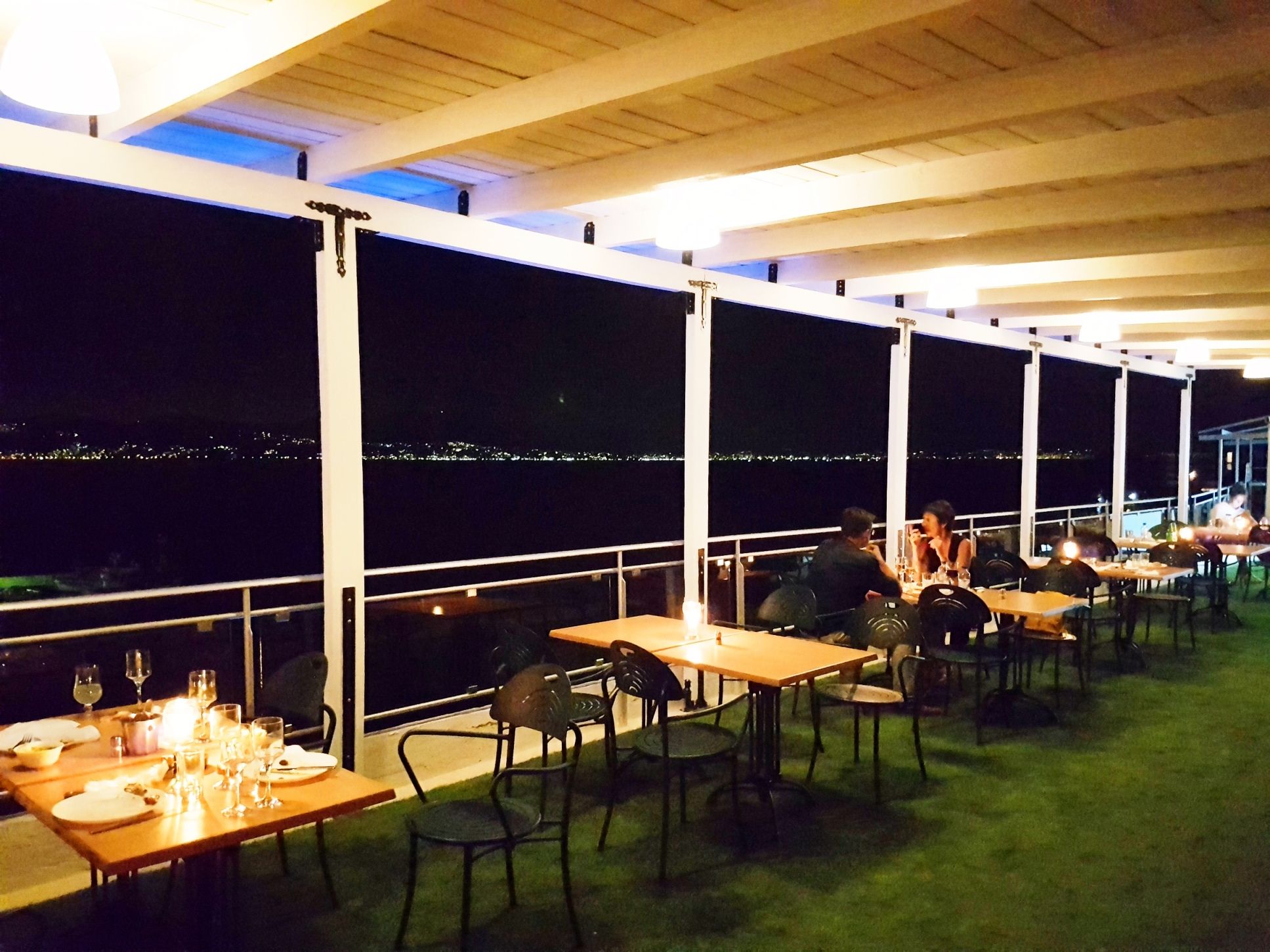 Uma área de refeições ao ar livre com mesas e cadeiras, vista da cidade iluminada à noite, e uma cobertura.