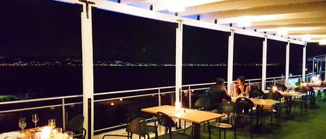 Uma área de refeições ao ar livre com mesas e cadeiras, vista da cidade iluminada à noite, e uma cobertura.