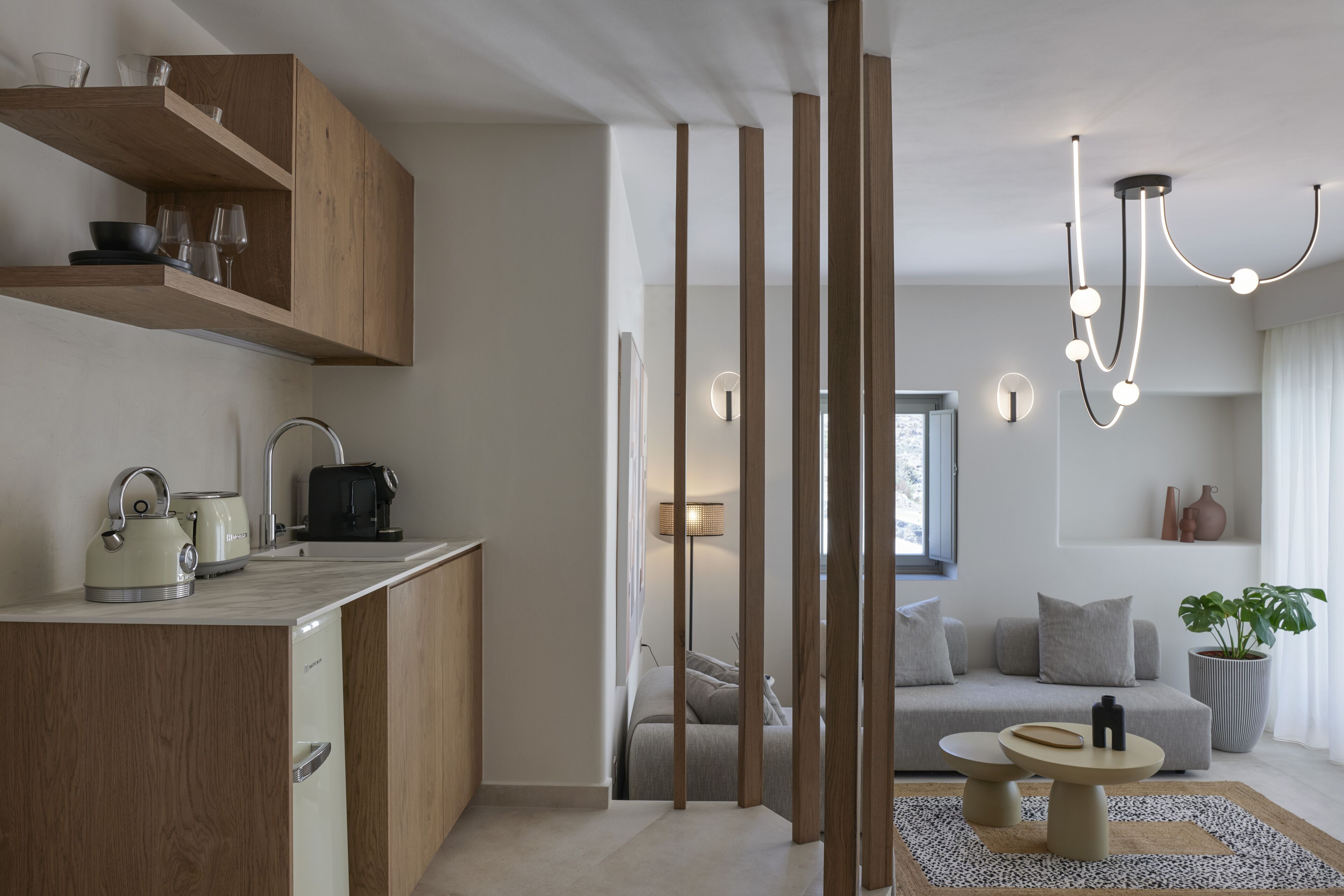 Cozinha moderna com armários de madeira, bancada com eletrodomésticos e vista para uma sala de estar com sofá e planta.