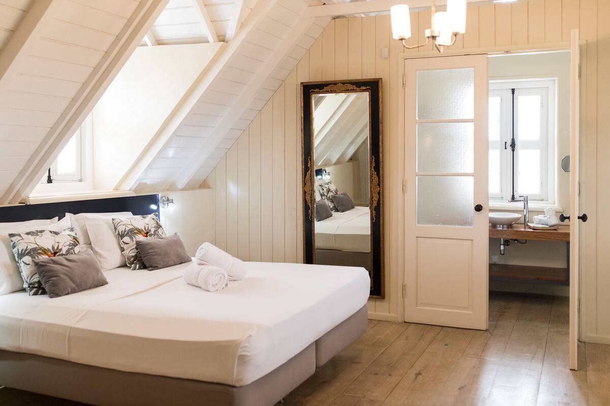 Quarto com uma cama grande, um espelho e um banheiro visível através de uma porta aberta.