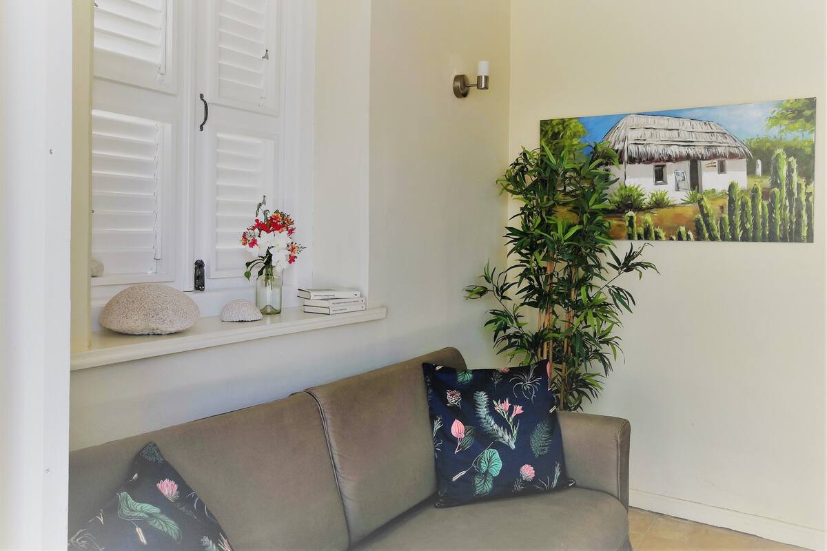 Uma sala de estar com sofá, janela com persianas, um vaso com flores, planta em vaso e um quadro na parede.