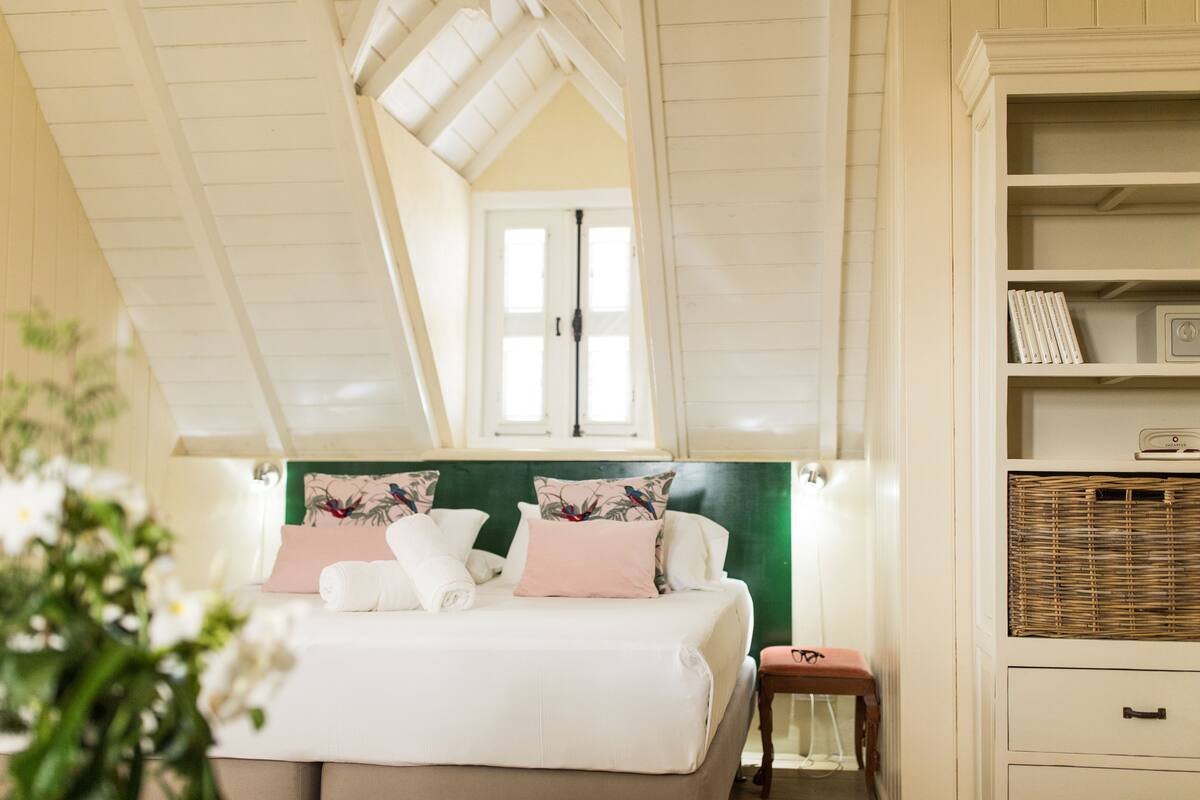 Quarto com teto alto, cama com almofadas florais, mesa de cabeceira e cesto de vime sobre um armário branco.