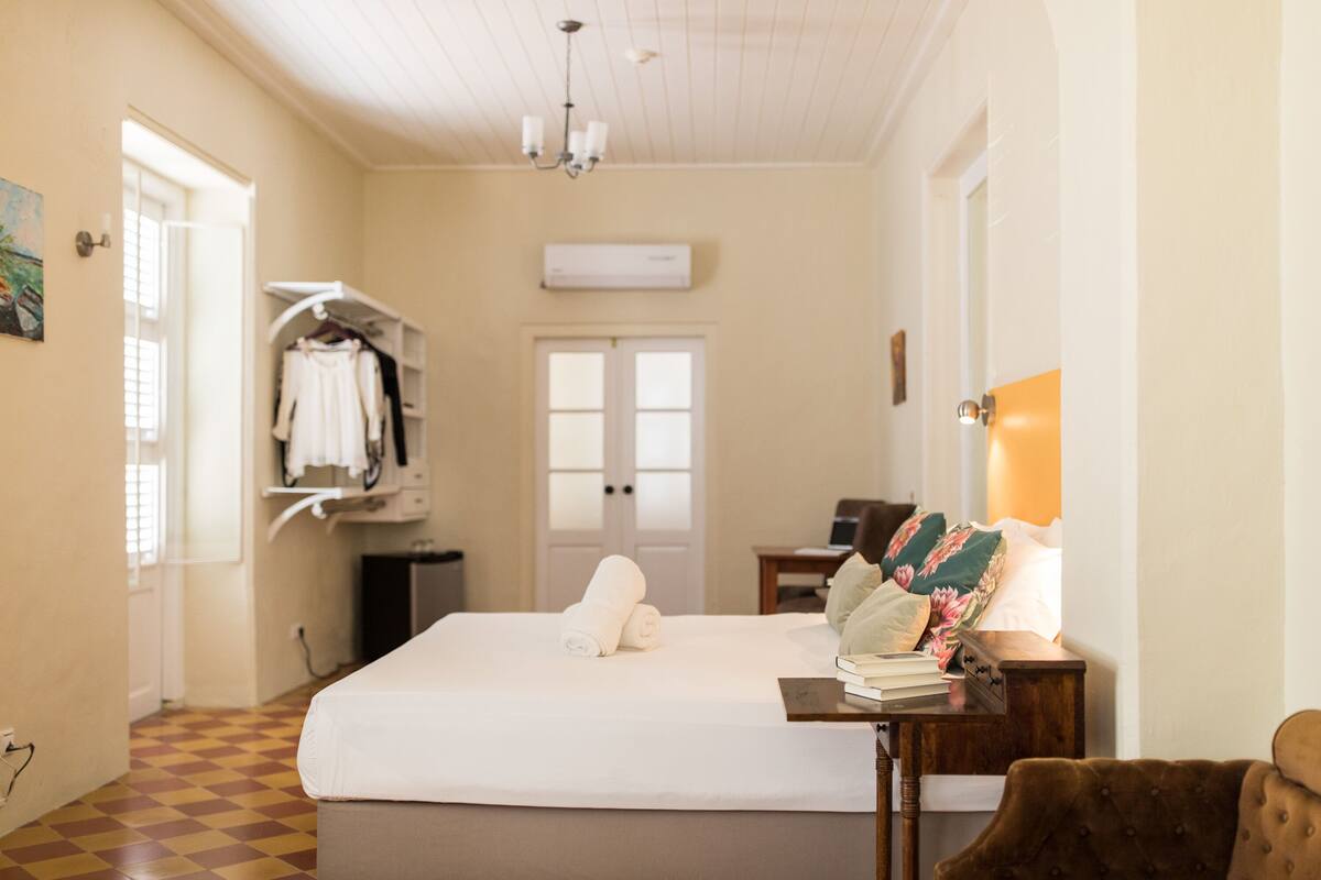 Quarto de hotel com uma cama, mesa de cabeceira com livros, luminária, guarda-roupa e ar condicionado.