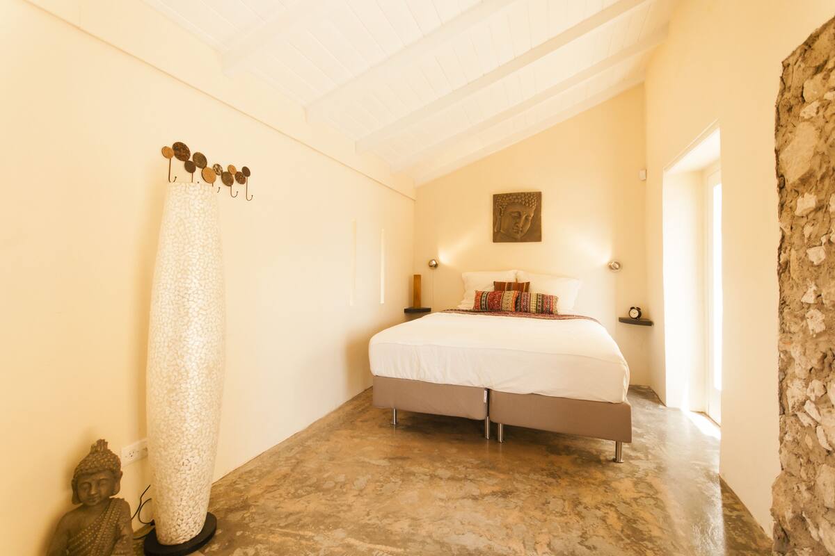 Quarto com cama, luminária na parede, estátua de Buda e um pilar texturizado.