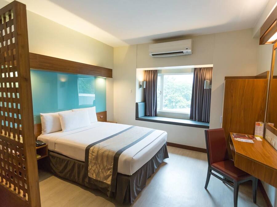 Quarto de hotel com cama, secretária, cadeira, janela com cortinas, unidade de ar condicionado e divisória de madeira.
