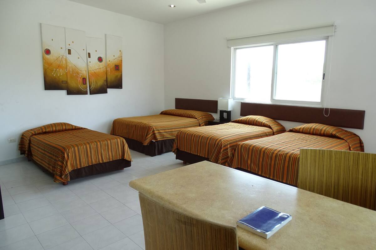 Quarto de hotel com três camas, uma mesa pequena e uma janela.