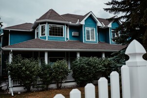 Uma casa de dois andares com varanda, cerca branca e uma grande árvore em primeiro plano.