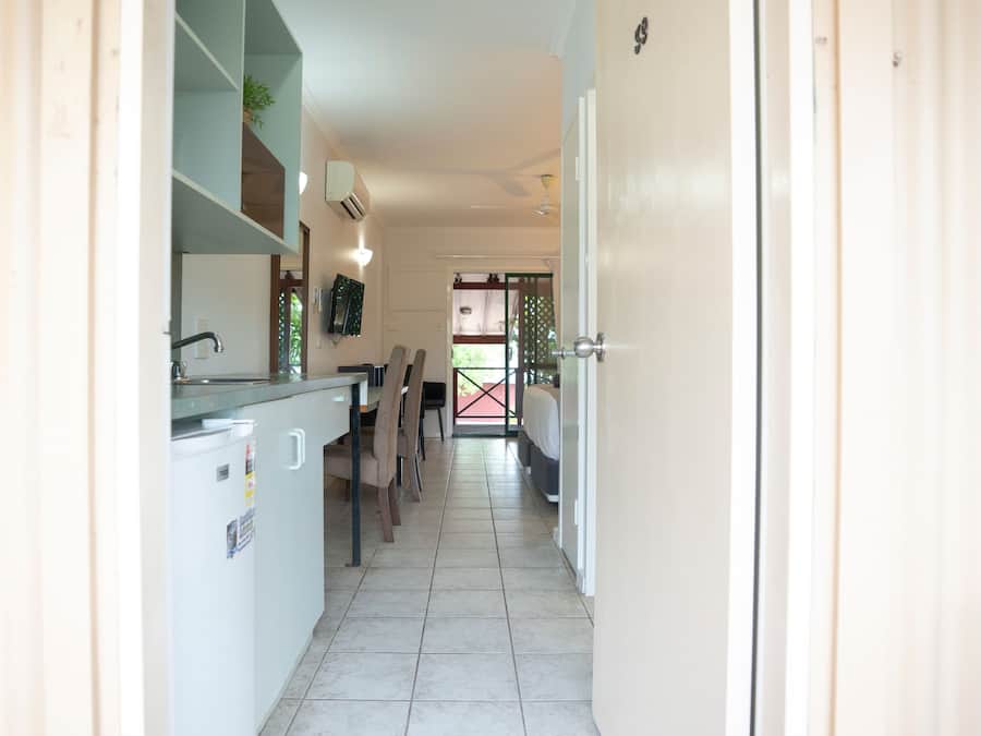 Corredor estreito com área de cozinha à esquerda, incluindo pia, armários e geladeira. O corredor possui piso de azulejos e uma porta à direita que dá para outro cômodo.