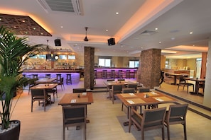 Restaurante com várias mesas e cadeiras, um espaço de bar e uma grande planta em vaso.