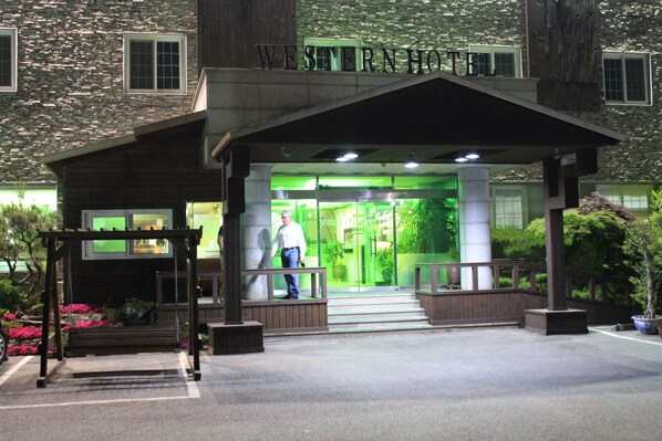 Entrada de hotel com varanda coberta, um homem caminhando em direção à entrada, e fachada de pedra.