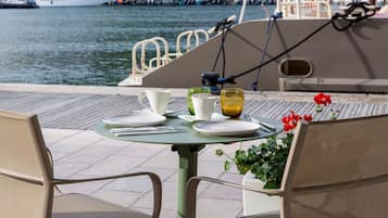 Mesa posta para dois, com pratos, copos e talheres, com vista para um areal marítimo com barcos.