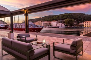 Terraço à beira-mar com mesas ao ar livre, vista para barcos e edifícios ao fundo.