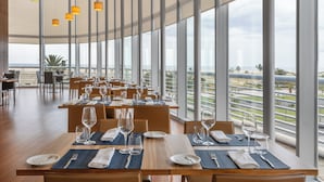Um restaurante com mesas preparadas para refeições, grandes janelas com vista para a praia e luminárias pendentes.