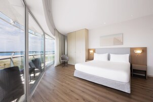 Quarto de hotel moderno com uma cama grande, uma cadeira, uma mesa de cabeceira e vista para o oceano através de janelas do chão ao teto.