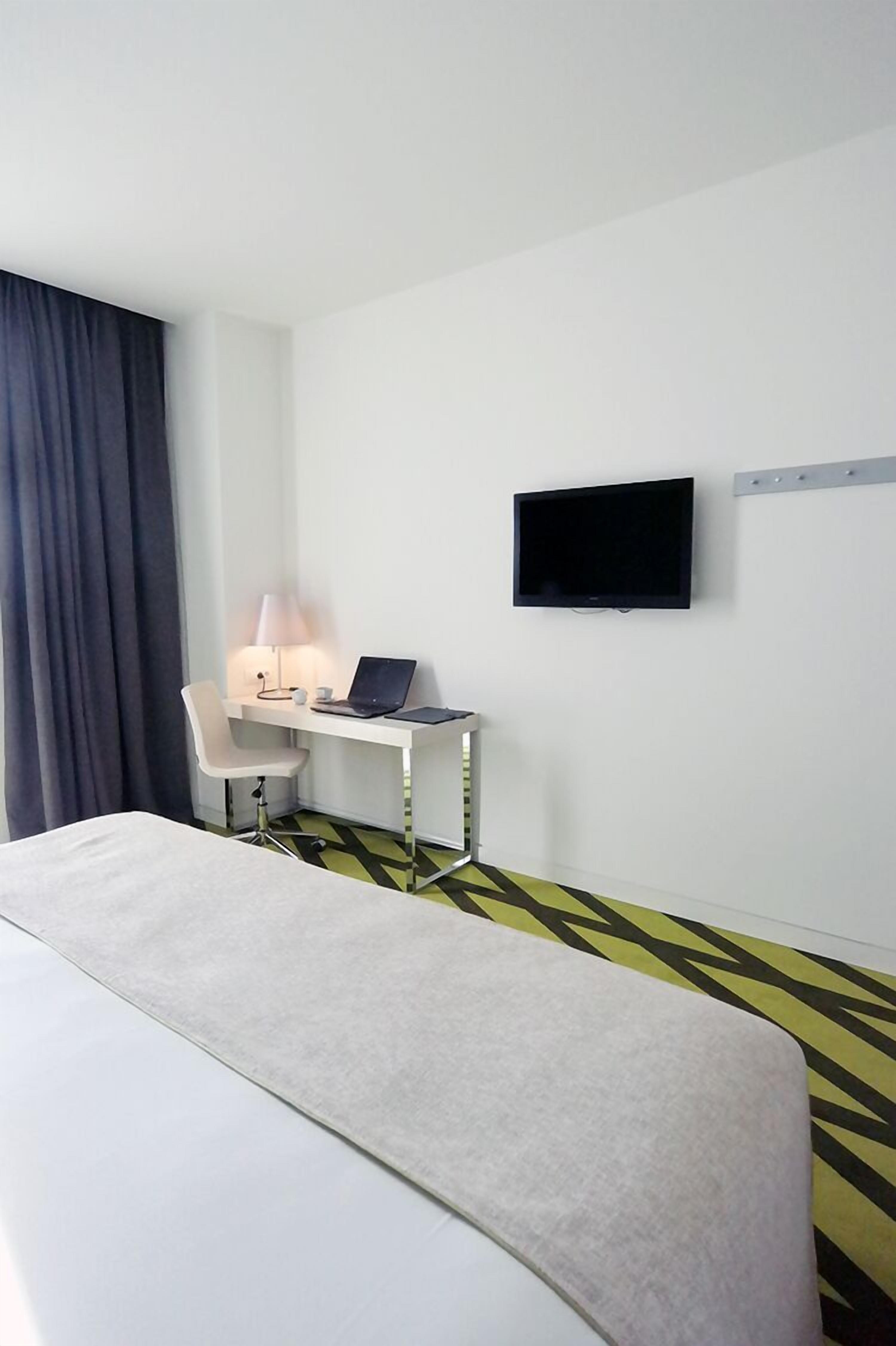 Quarto de hotel com cama, escrivaninha com laptop, cadeira, TV fixada na parede e tapete estampado.
