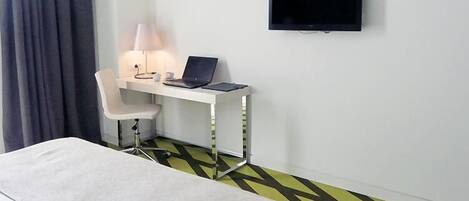 Quarto de hotel com cama, escrivaninha com laptop, cadeira, TV fixada na parede e tapete estampado.