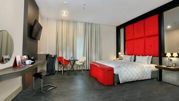 Quarto de hotel moderno com uma cama grande, uma escrivaninha com cadeira, uma TV e uma parede de destaque vermelha.
