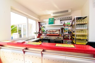 Uma área de bar com balcão vermelho, prateleiras com garrafas e copos, uma máquina de café e um quadro de cardápio.