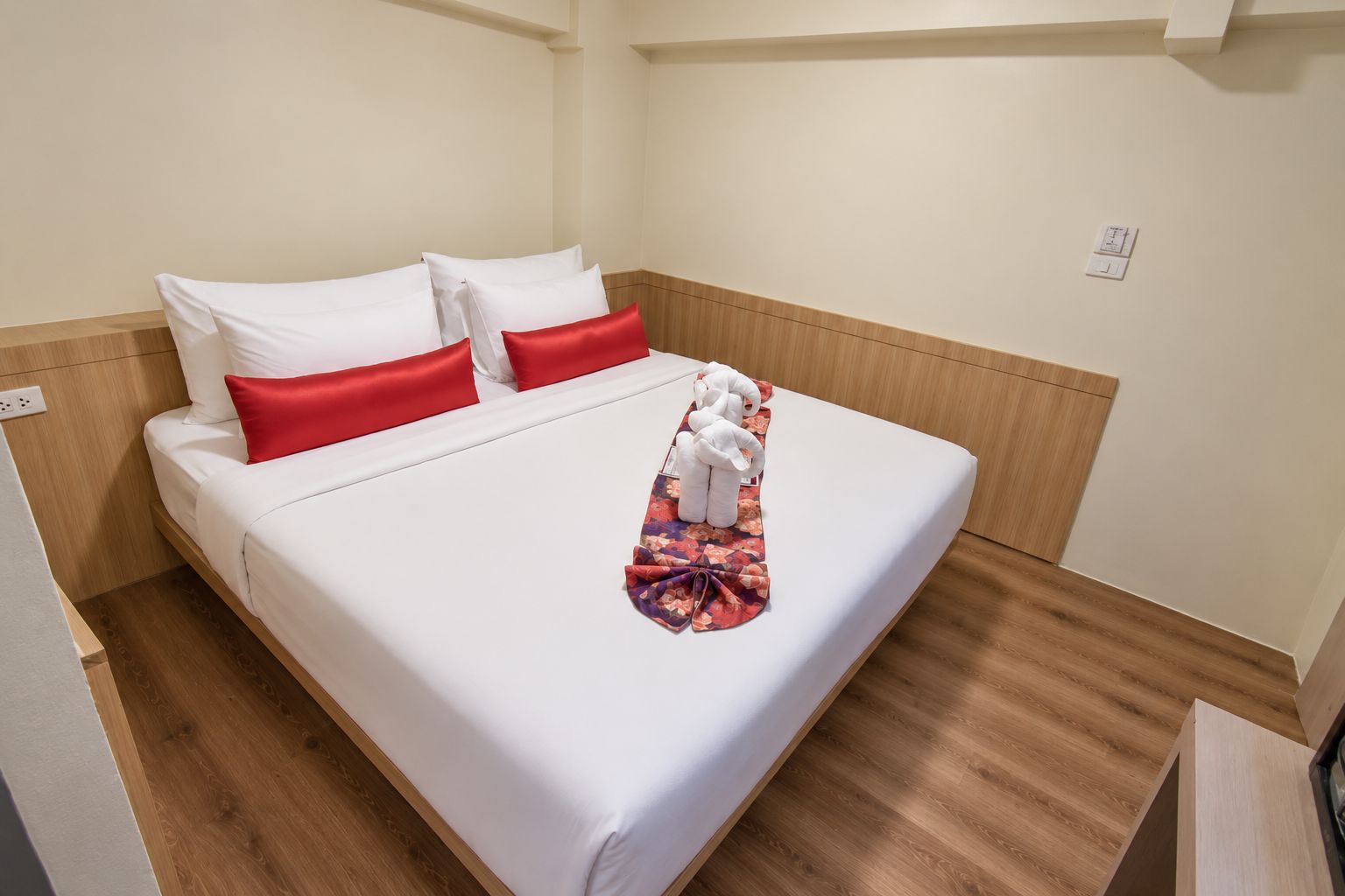 Quarto de hotel com cama, roupa de cama branca, almofadas vermelhas e toalha florida sobre a cama.