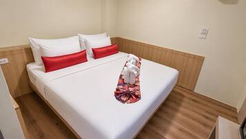 Quarto de hotel com cama, roupa de cama branca, almofadas vermelhas e toalha florida sobre a cama.