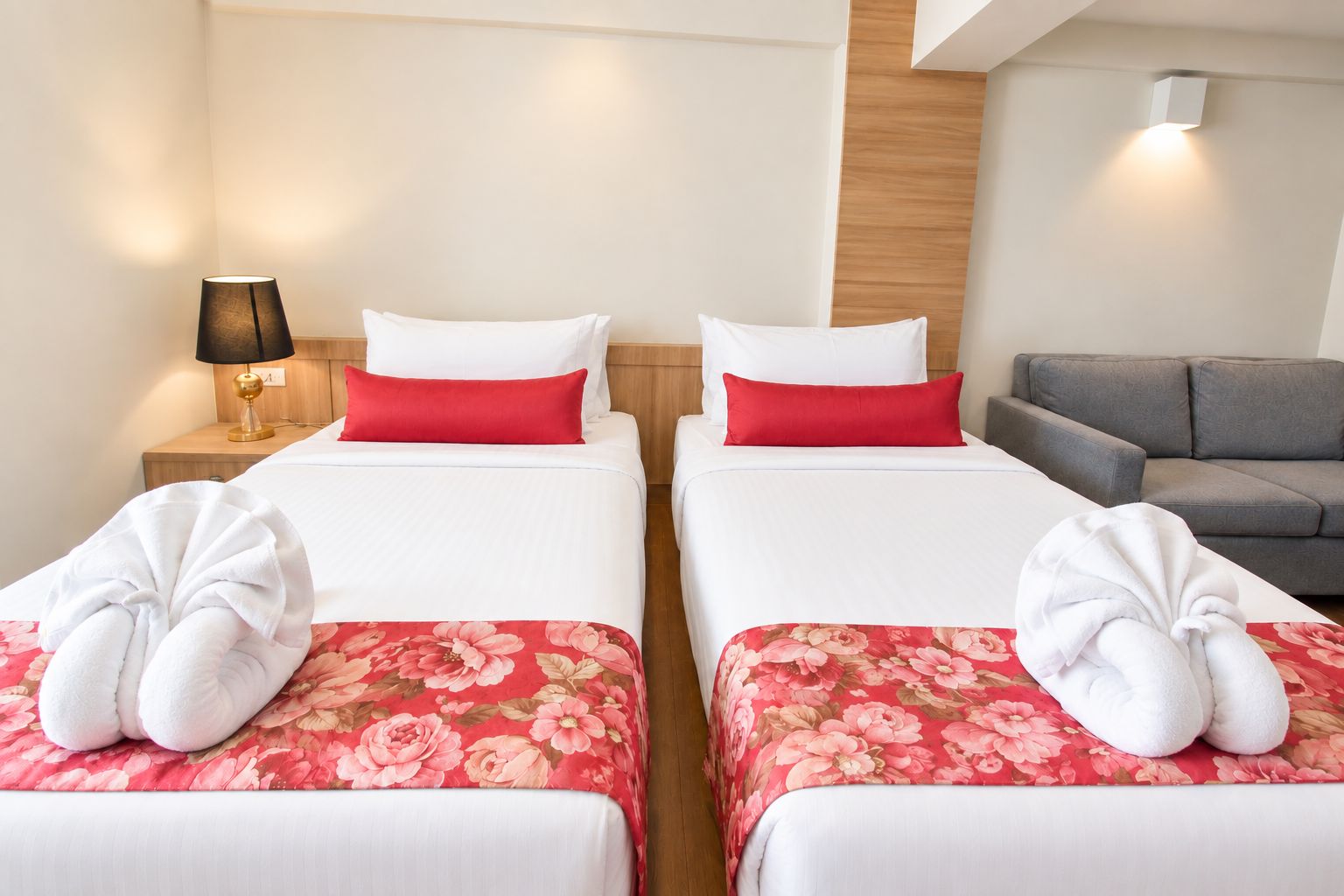 Quarto de hotel com duas camas, cada uma com colcha florida vermelha e almofadas brancas. Há uma mesinha de cabeceira com abajur e uma pequena mesa com um aviso. Um sofá cinza é visível ao fundo.