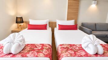 Quarto de hotel com duas camas, cada uma com colcha florida vermelha e almofadas brancas. Há uma mesinha de cabeceira com abajur e uma pequena mesa com um aviso. Um sofá cinza é visível ao fundo.