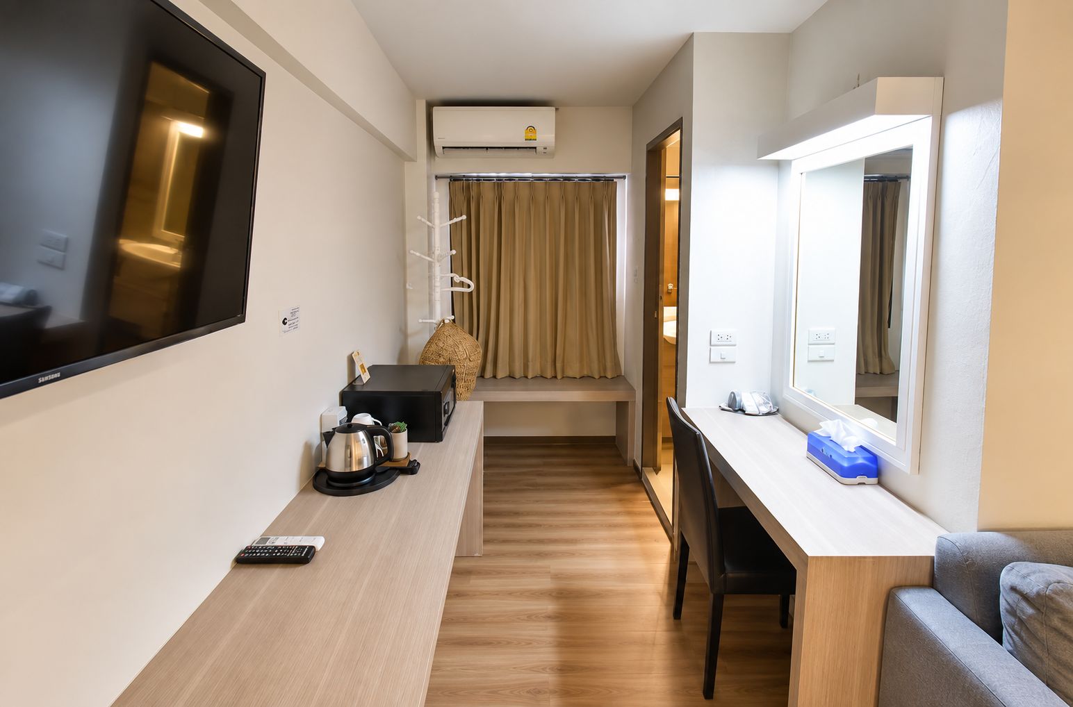 Quarto de hotel compacto com TV de tela plana, escrivaninha com cadeira, pequena cozinha e área de estar com sofá.