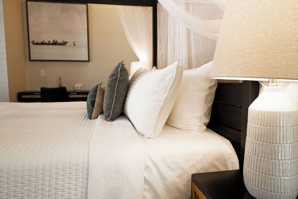 Cama bem arrumada com roupa de cama branca e várias almofadas, mesa de cabeceira com abajur e um quadro na parede.