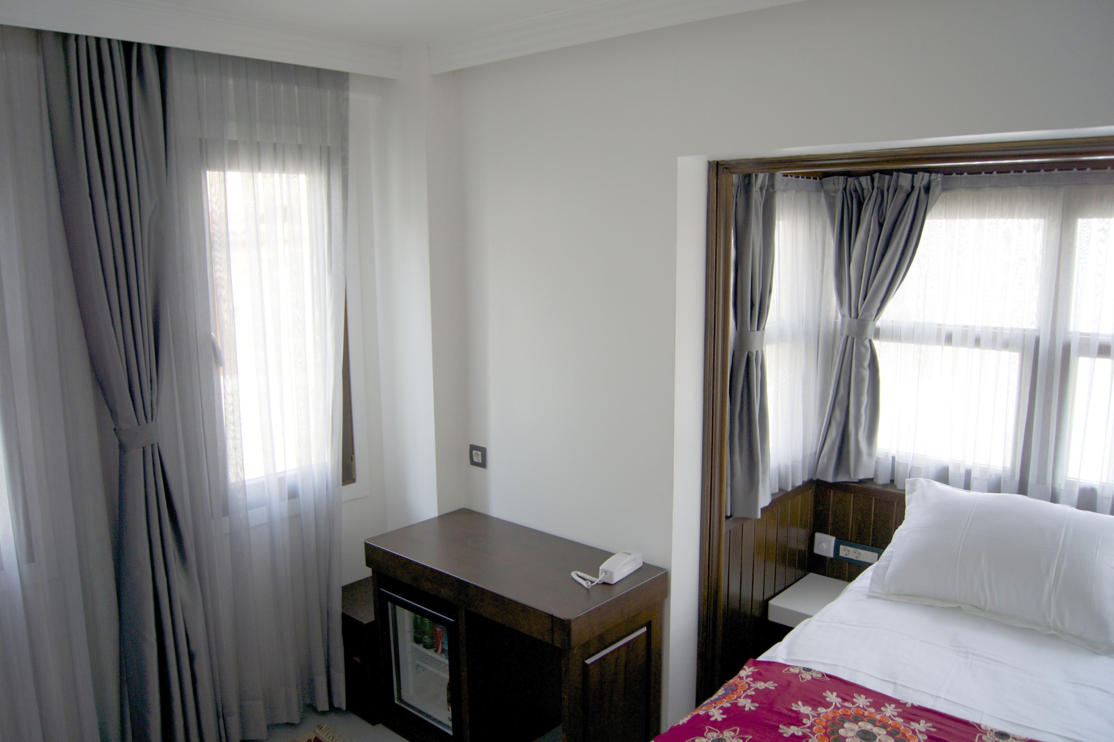 Quarto de hotel com cama, escrivaninha com telefone, mini-geladeira e cortinas.
