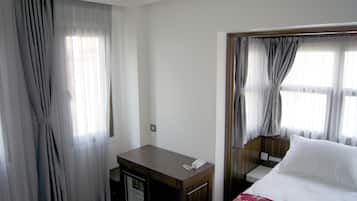 Quarto de hotel com cama, escrivaninha com telefone, mini-geladeira e cortinas.