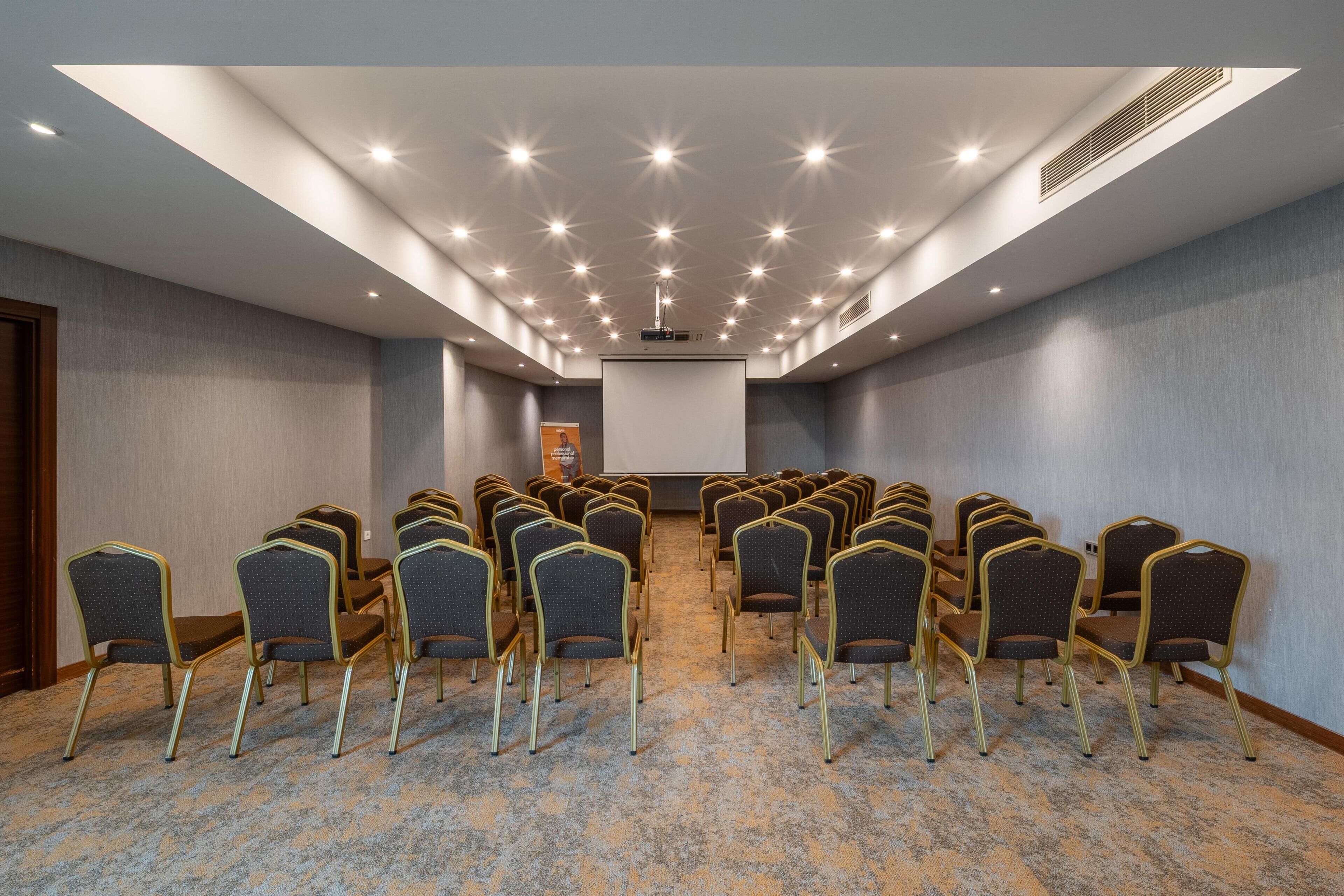 Uma sala de conferência com fileiras de cadeiras, uma tela de projetor e luminárias no teto.