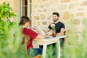 Um casal sentado à mesa com um muro de pedra ao fundo, uma planta em vaso e um croissant.